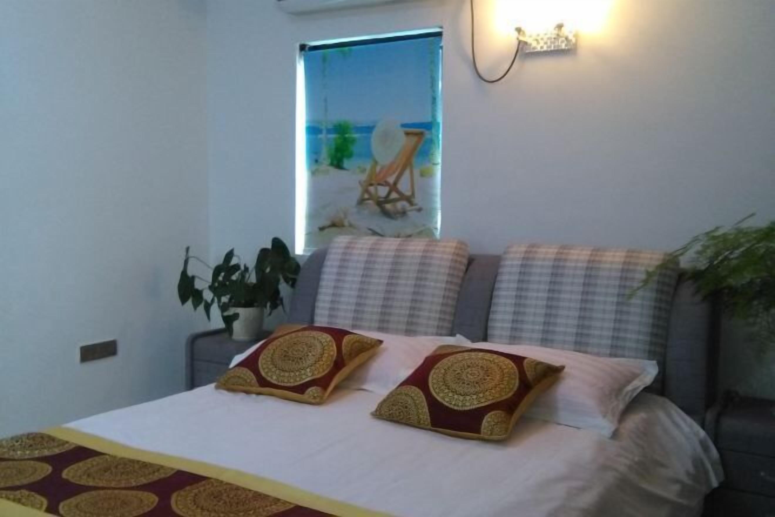 Quarto com cama, almofadas, mesa de cabeceira, planta em vaso, janela com vista para a praia e luminária fixada na parede.