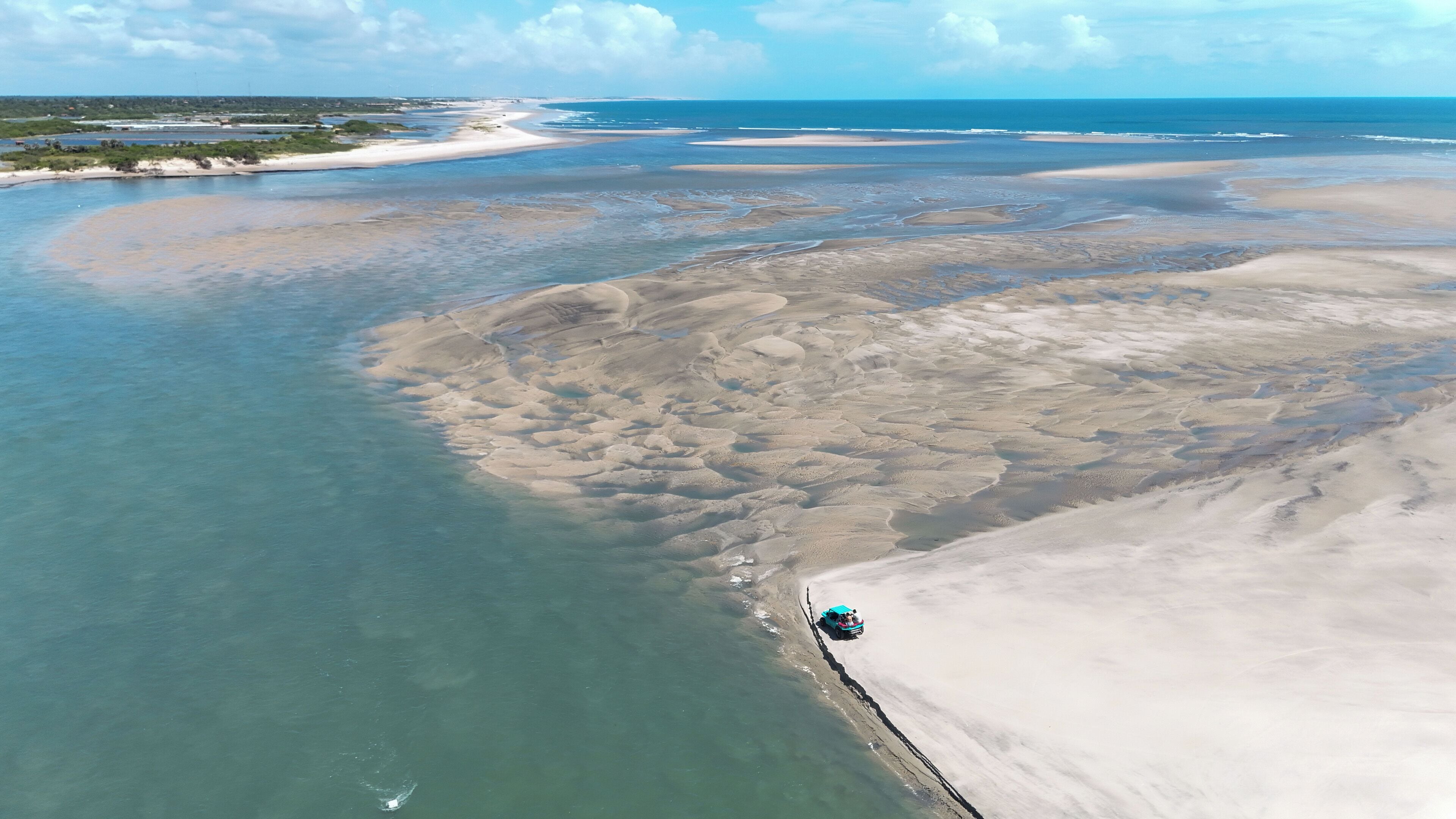 Vista aérea de uma área costeira com uma praia arenosa, um barco pequeno e uma linha de costa distante com vegetação e edifícios.