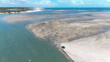 Vista aérea de uma área costeira com uma praia arenosa, um barco pequeno e uma linha de costa distante com vegetação e edifícios.