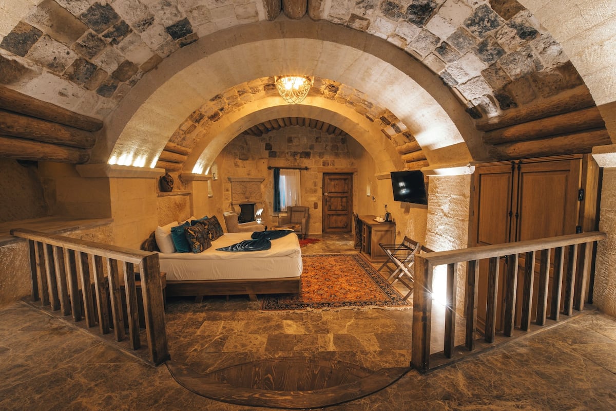 Um quarto com soalho de pedra, cama com almofadas, balaústres de madeira e um arco de pedra.