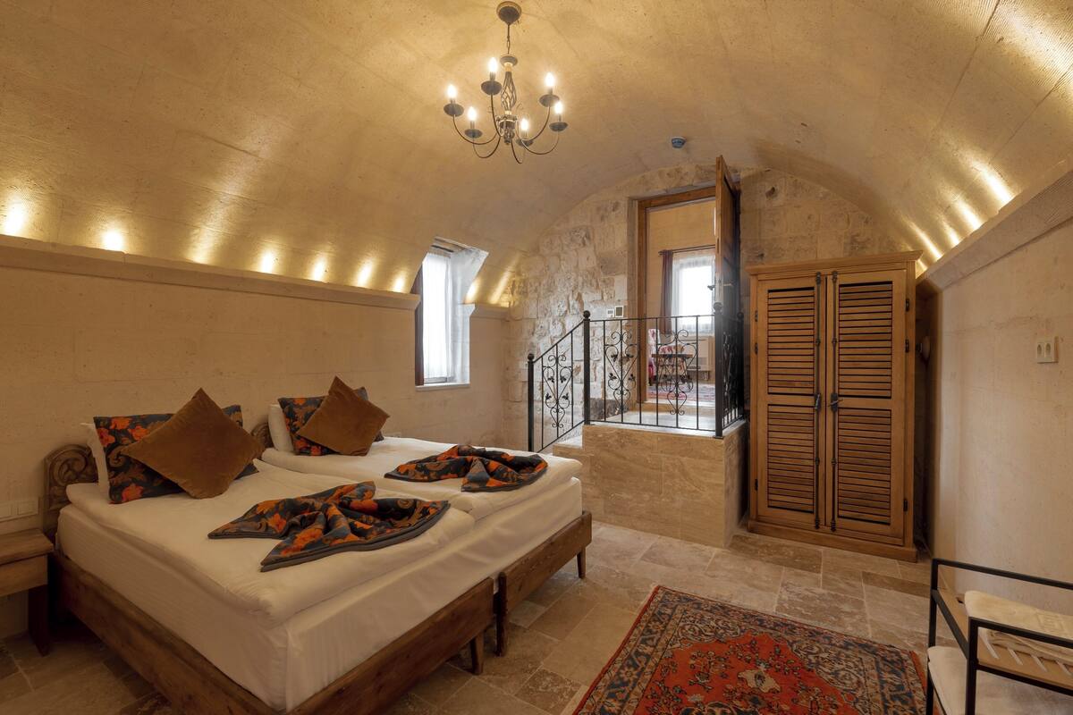 Quarto com duas camas, um candelabro, um guarda-roupa de madeira e vista para o exterior.