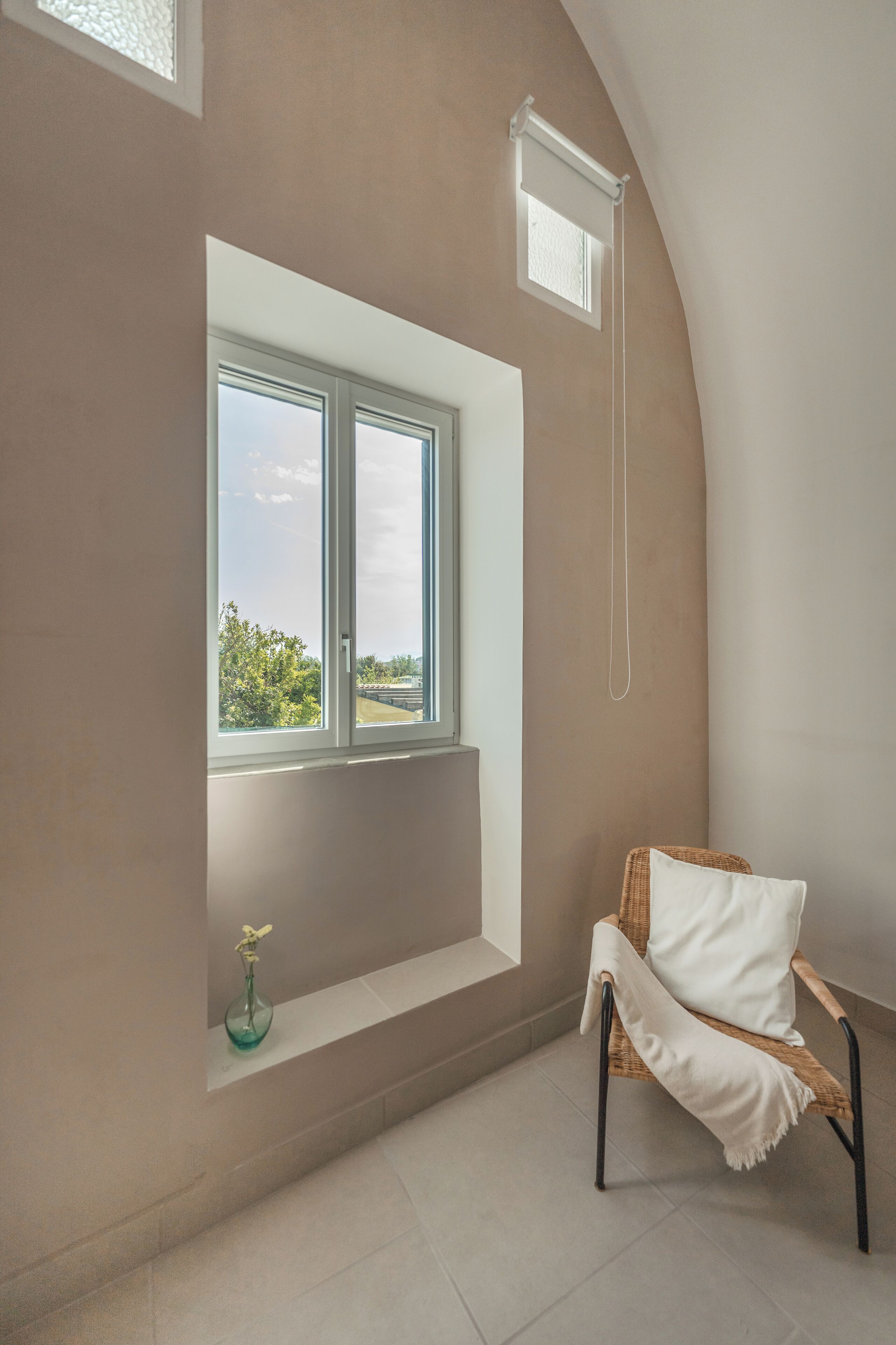 Um quarto com uma janela grande, uma cadeira de vime com almofada branca e um pequeno vaso no parapeito.