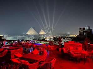 Um bar no terraço com mobiliário de vime e iluminação vermelha, com vista para um horizonte urbano onde se destacam duas pirâmides iluminadas.