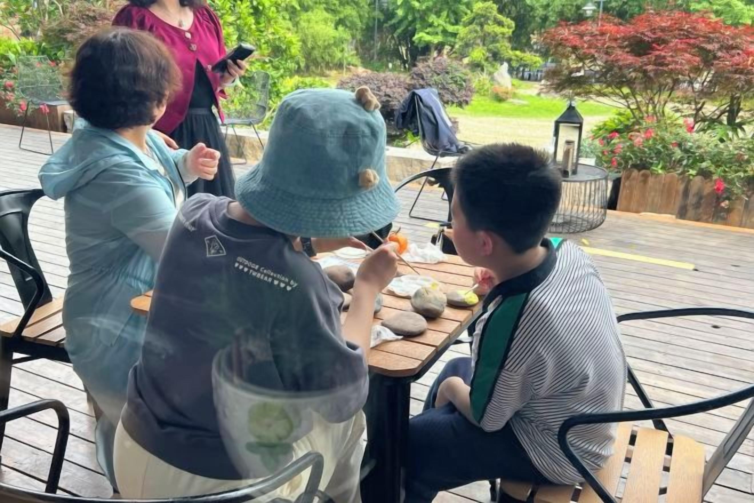 Um grupo de pessoas sentadas em uma mesa de madeira ao ar livre, com cadeiras, em um ambiente com vegetação e uma cerca ao fundo.