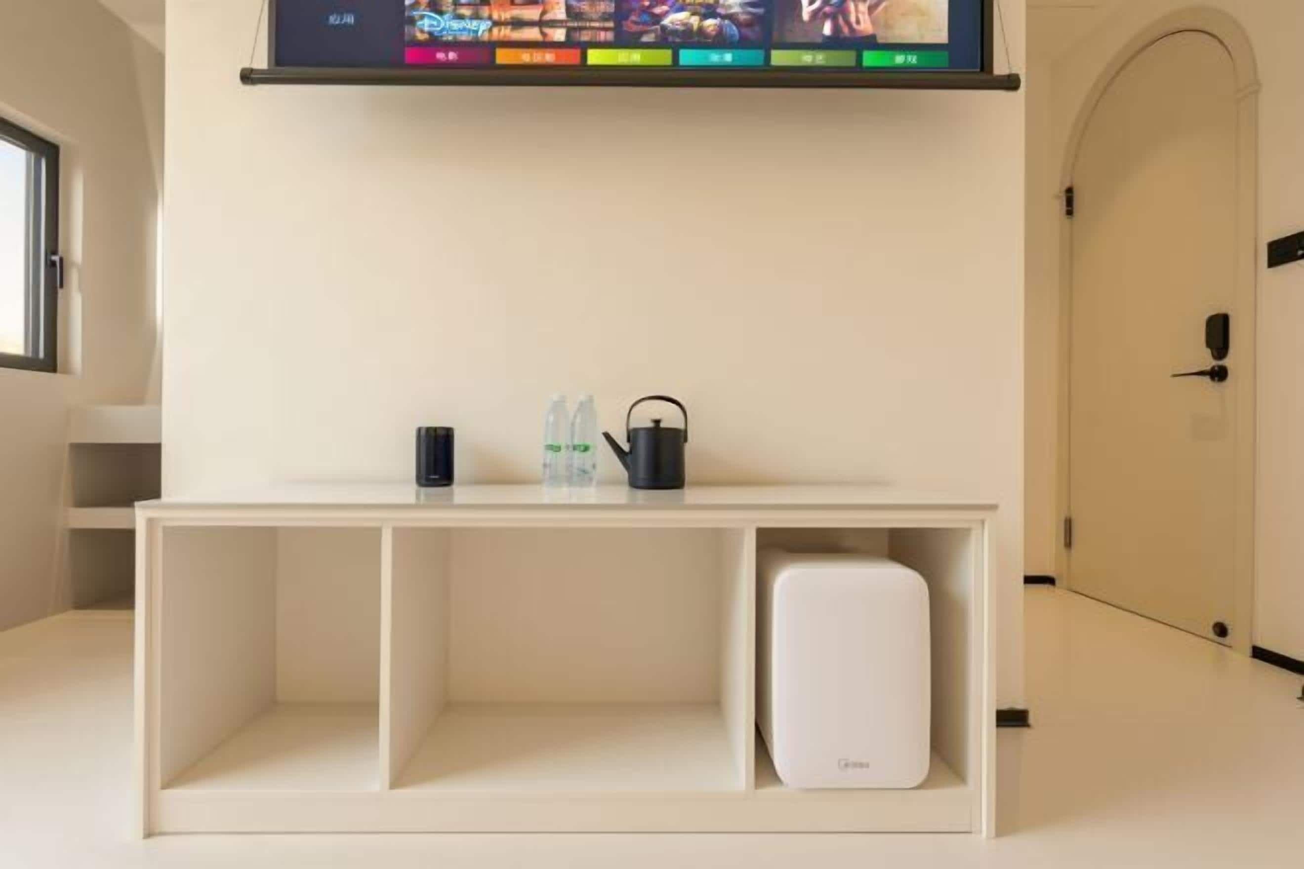 Quarto de hotel com uma mesa de consola branca, uma televisão de ecrã plano montada acima e um mini frigorífico branco.