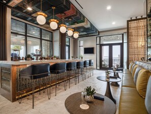 Uma área de bar moderna com um balcão comprido, vários banquinhos e um espaço de estar com um sofá e uma mesa redonda.
