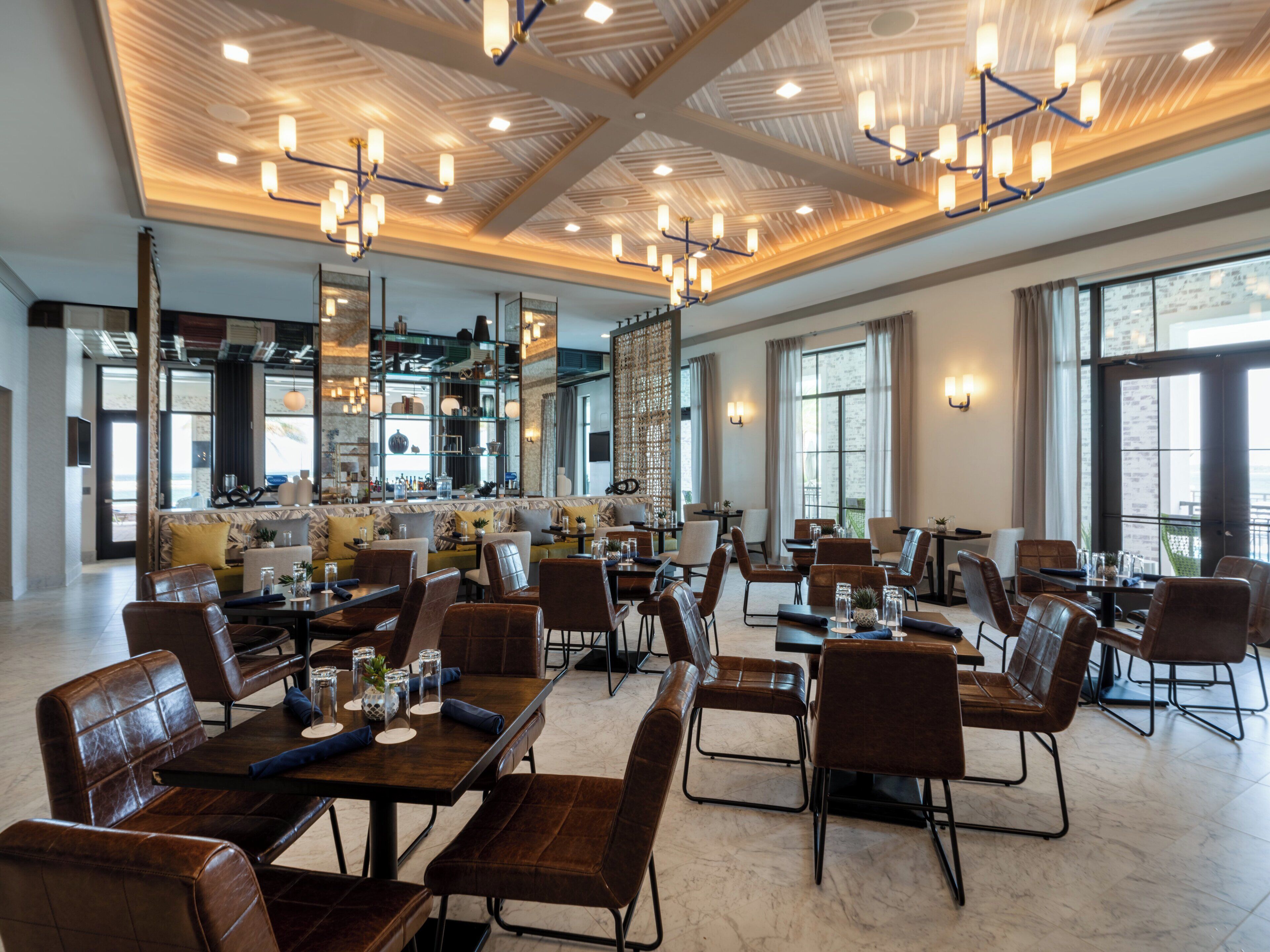 Uma sala de jantar ampla, com várias mesas e cadeiras, teto alto com luminárias modernas e grandes janelas que proporcionam luz natural.