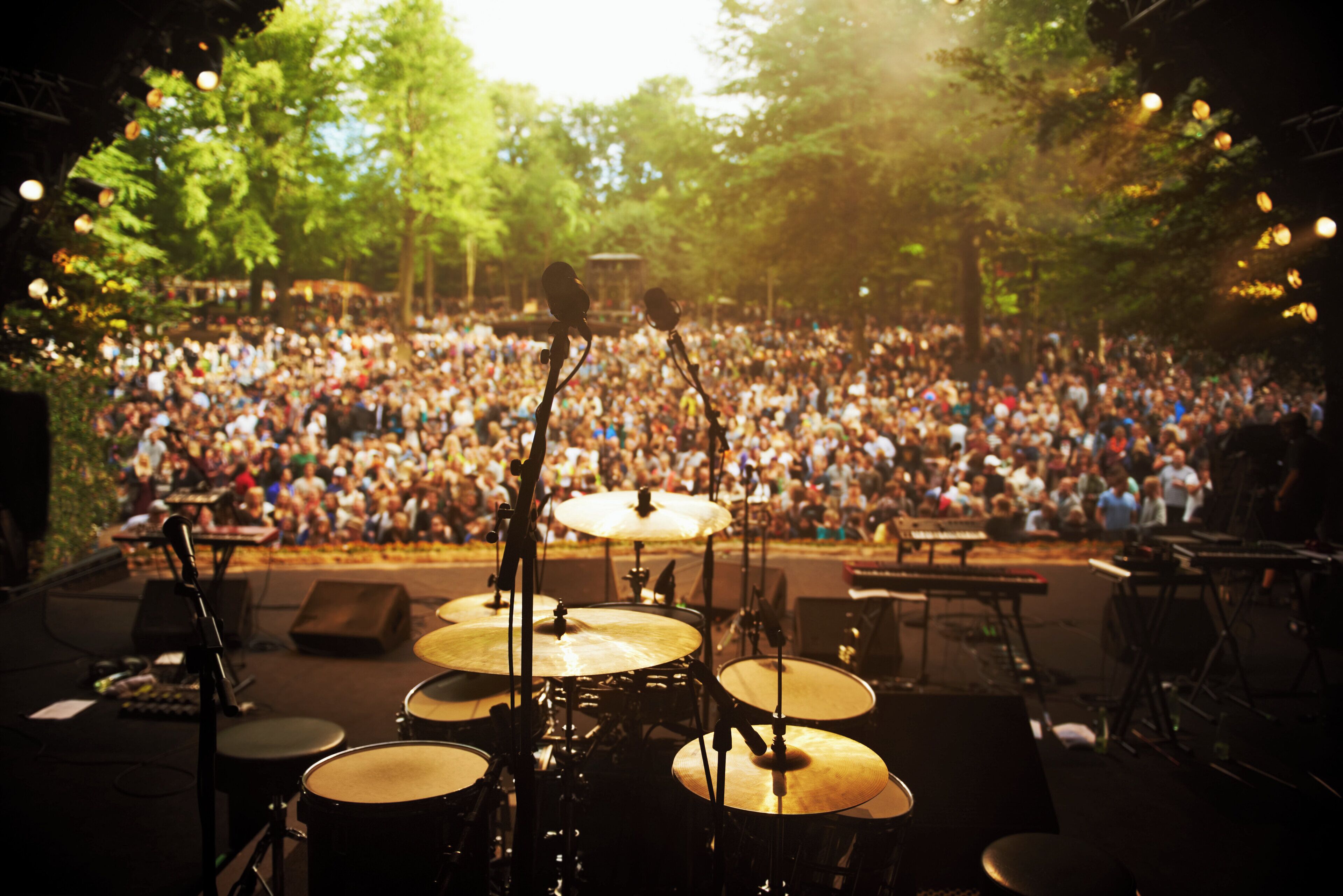 Palco de concerto ao ar livre com uma bateria em primeiro plano e uma grande multidão ao fundo.