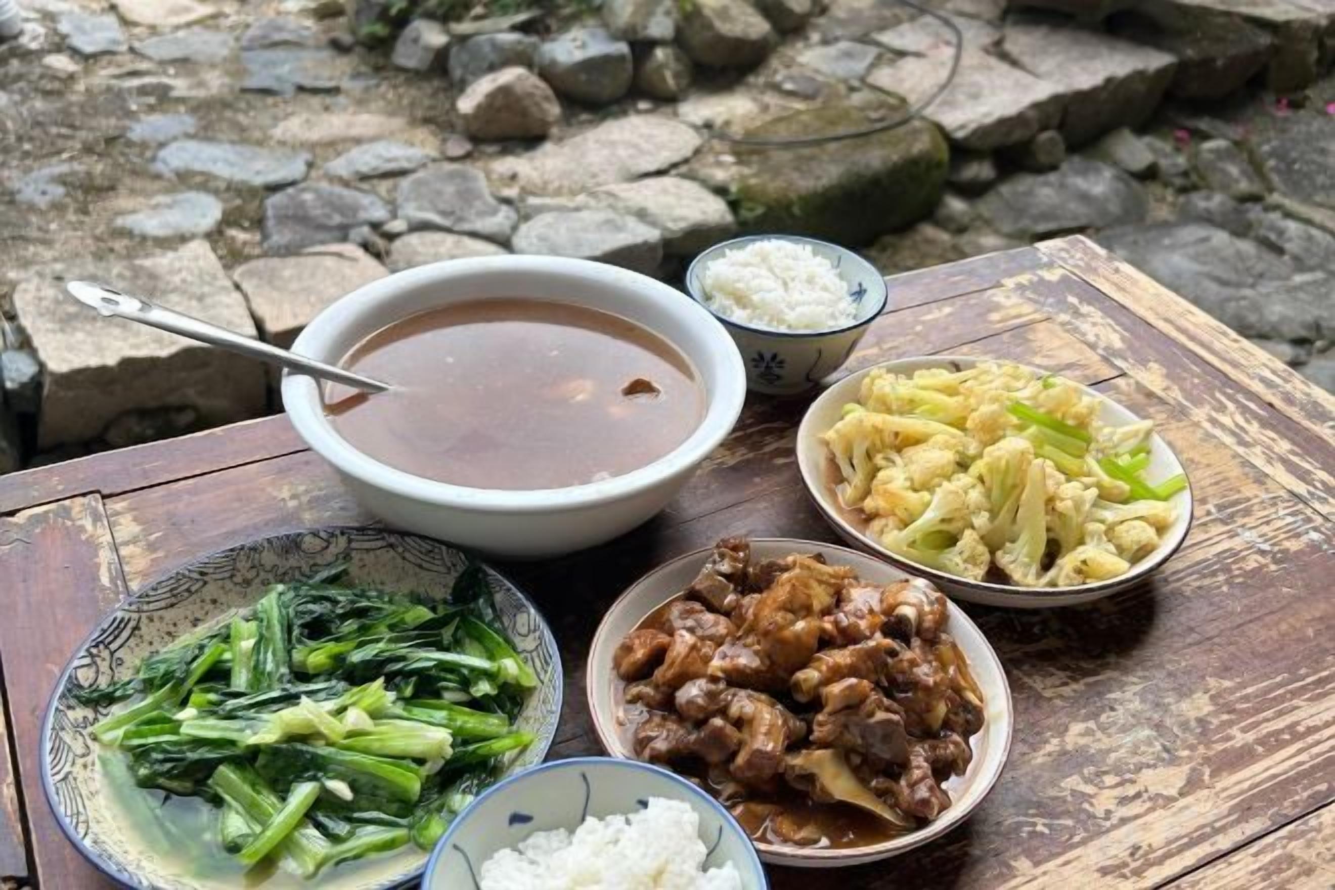 Uma mesa de madeira com diversos pratos, incluindo uma tigela de sopa, um prato de legumes, uma tigela de arroz e um prato com carne.