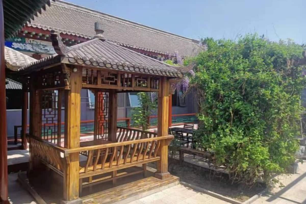 Um pavilhão de madeira tradicional com telhado de telhas, rodeado por vegetação e um edifício com telhado de telhas ao fundo.