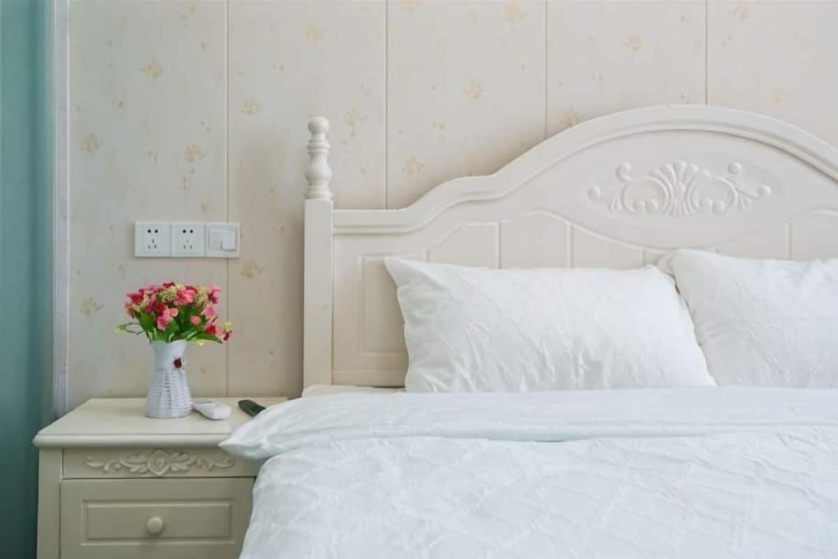Uma cama com roupa de cama branca, uma mesa de cabeceira com um vaso de flores e uma parede com papel de parede floral.