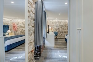 Quarto com parede de pedra, cama com cabeceira azul, escrivaninha com abajur e uma cadeira.