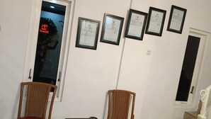 Quarto com duas cadeiras de vime com almofadas vermelhas, uma pequena mesa de madeira e uma escada. Há diplomas emoldurados na parede.