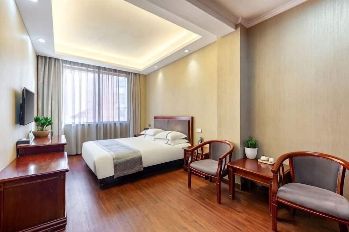Quarto de hotel com cama, duas cadeiras, escrivaninha, televisão e janela com cortinas.