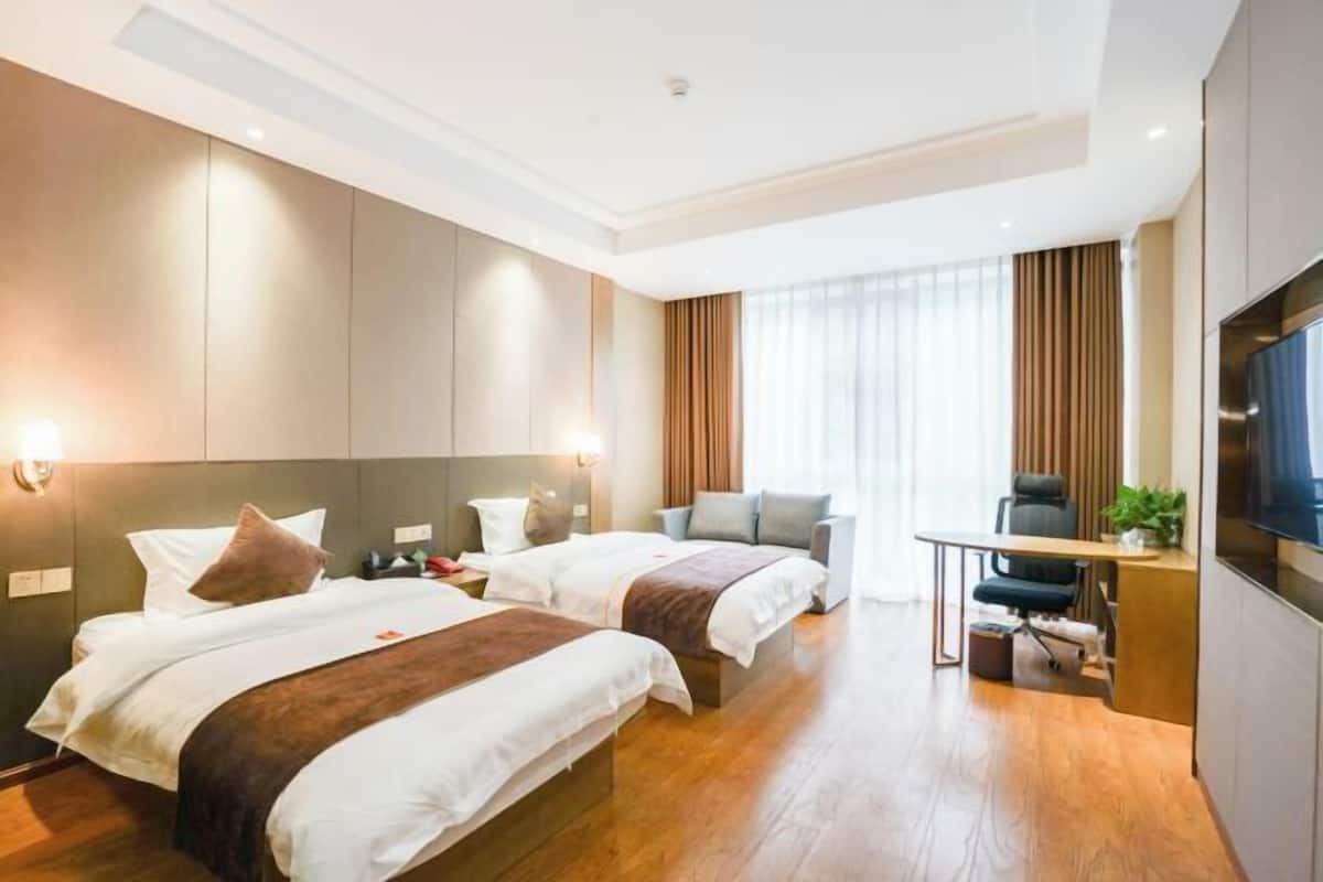 Quarto de hotel com duas camas, uma mesa, uma cadeira, uma TV e uma janela grande com cortinas.