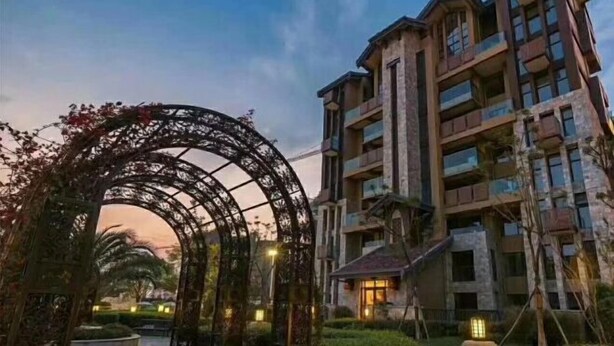 Um edifício residencial com vários andares, varandas, área de jardim com um arco decorativo e uma entrada bem iluminada.
