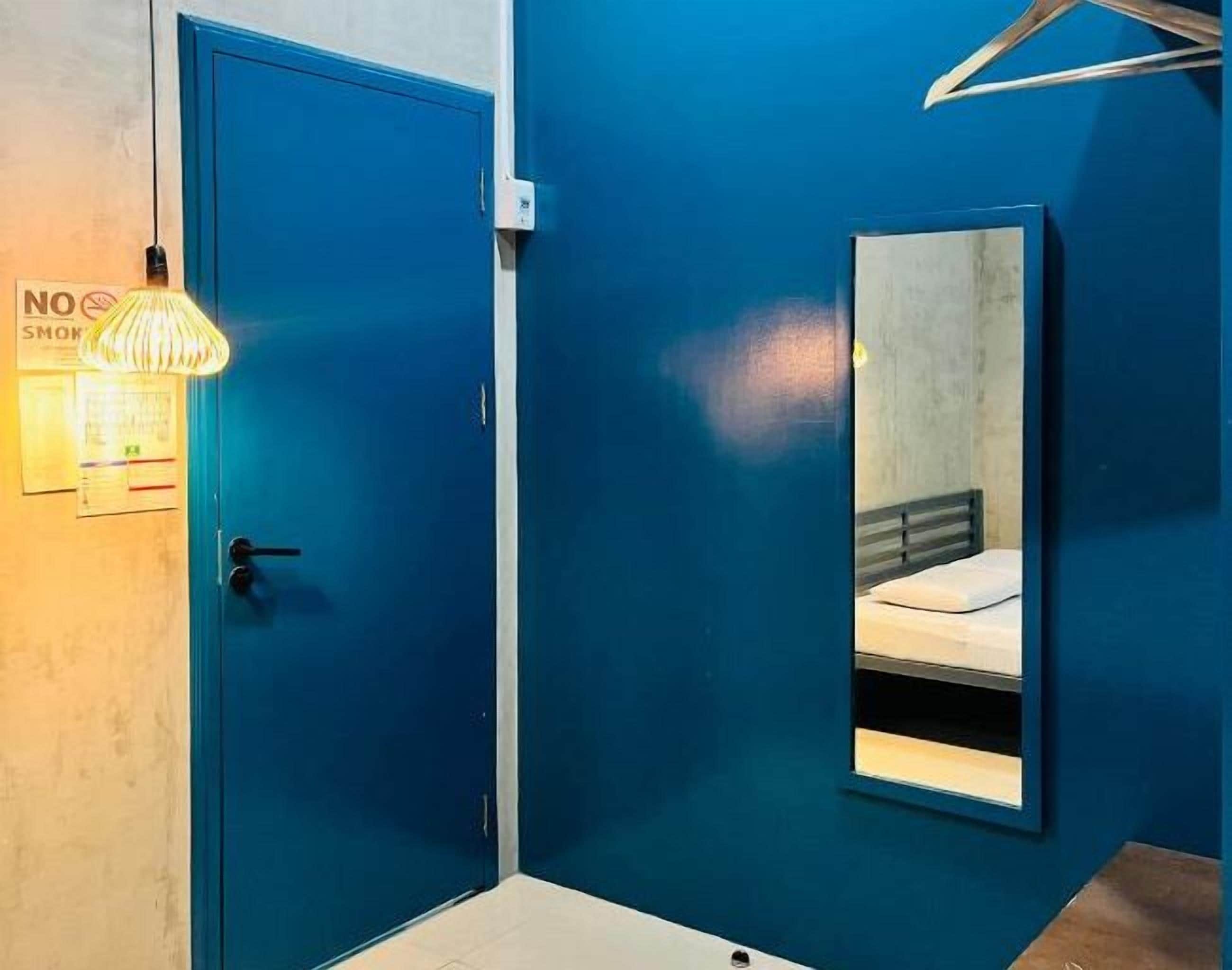 Quarto com porta azul, luminária pendente e espelho refletindo uma cama.