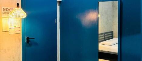 Quarto com porta azul, luminária pendente e espelho refletindo uma cama.