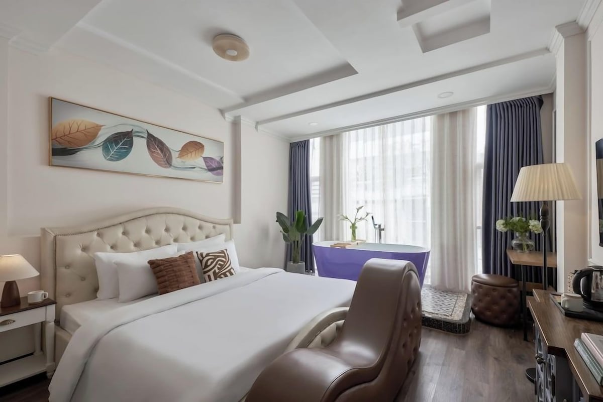 Quarto de hotel com uma cama grande, uma cadeira, uma escrivaninha, uma janela com cortinas e um quadro na parede.