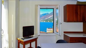 Quarto com cama, escrivaninha com televisão, janela com vista para montanhas e mar, e armários de madeira.
