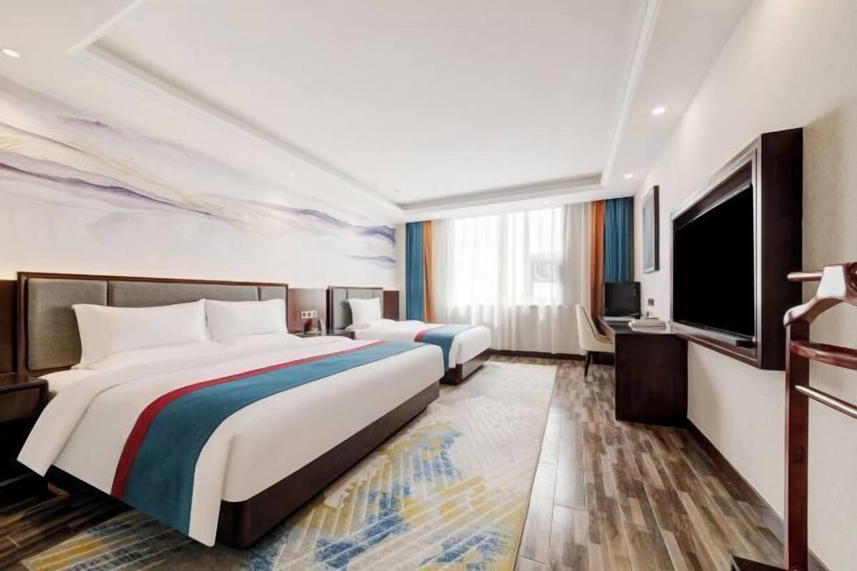 Quarto de hotel com uma cama grande, uma cama menor, uma mesa com televisão de tela plana e uma janela ampla com cortinas.