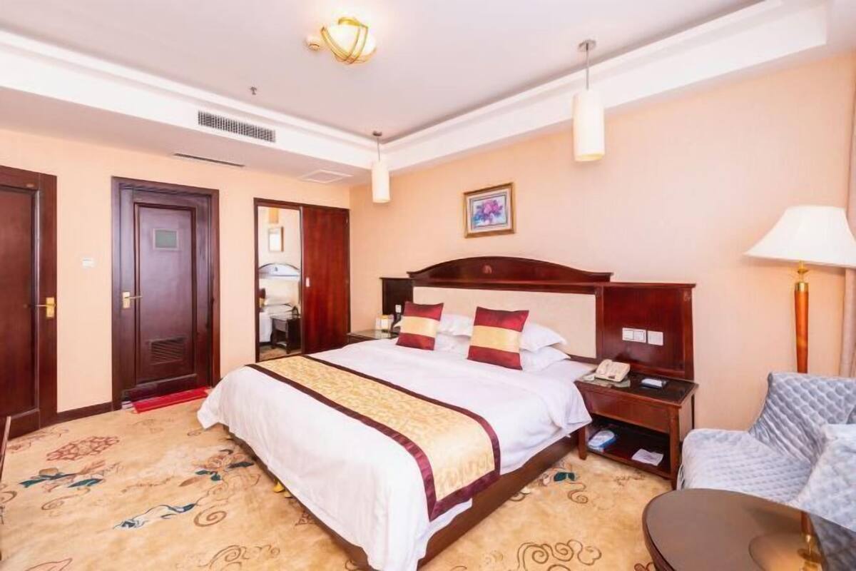 Quarto de hotel com uma cama grande, uma mesa de cabeceira com abajur, uma cadeira e piso coberto por carpete.