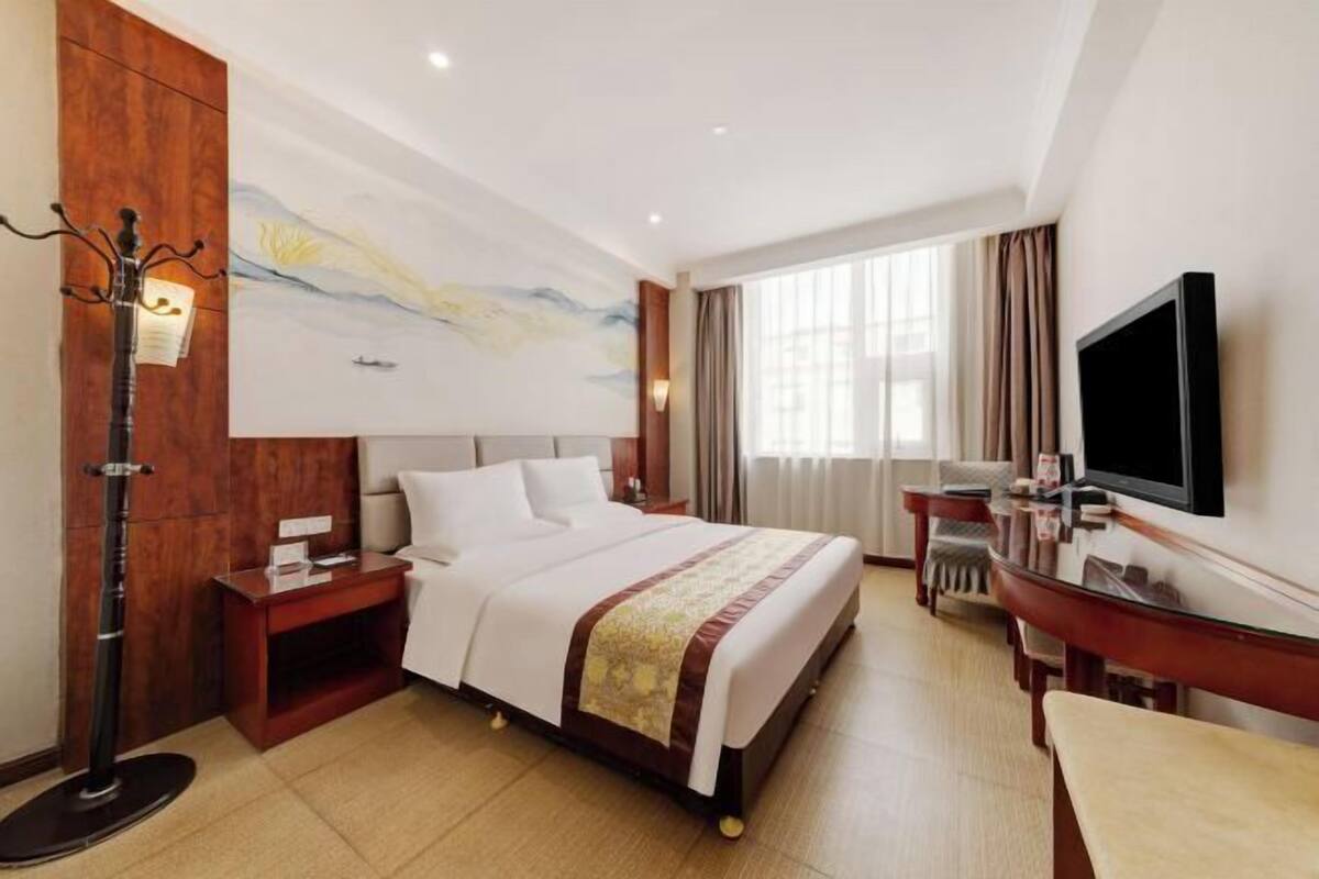 Quarto de hotel com uma cama grande, uma escrivaninha, televisão de tela plana, cabideiro e um quadro na parede.