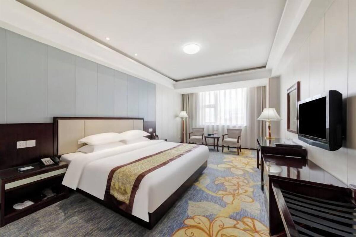 Quarto de hotel com uma cama grande, televisão, escrivaninha com luminária e uma mesa pequena.