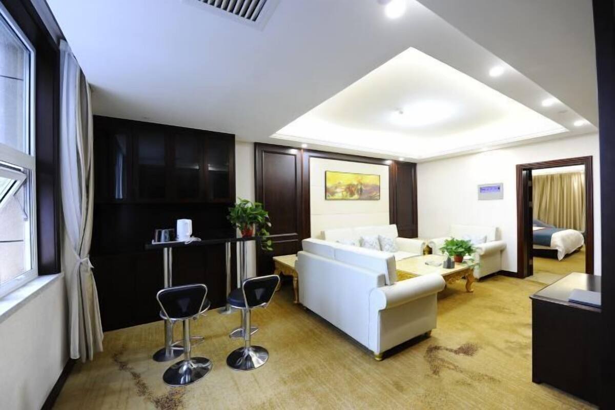 Quarto de hotel moderno com área de estar, mesa de jantar com banquetas, sofá, cama e uma pequena cozinha.