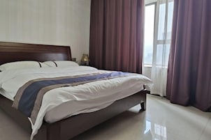 Uma cama bem arrumada com cabeceira de madeira, abajur e vista para o exterior através de grandes janelas com cortinas.