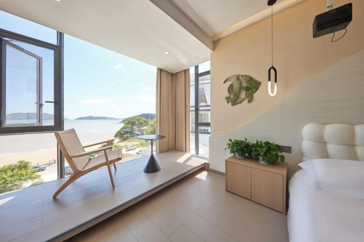 Quarto de hotel moderno com uma janela grande, uma cama, uma mesa de cabeceira com uma planta e uma cadeira perto da janela.