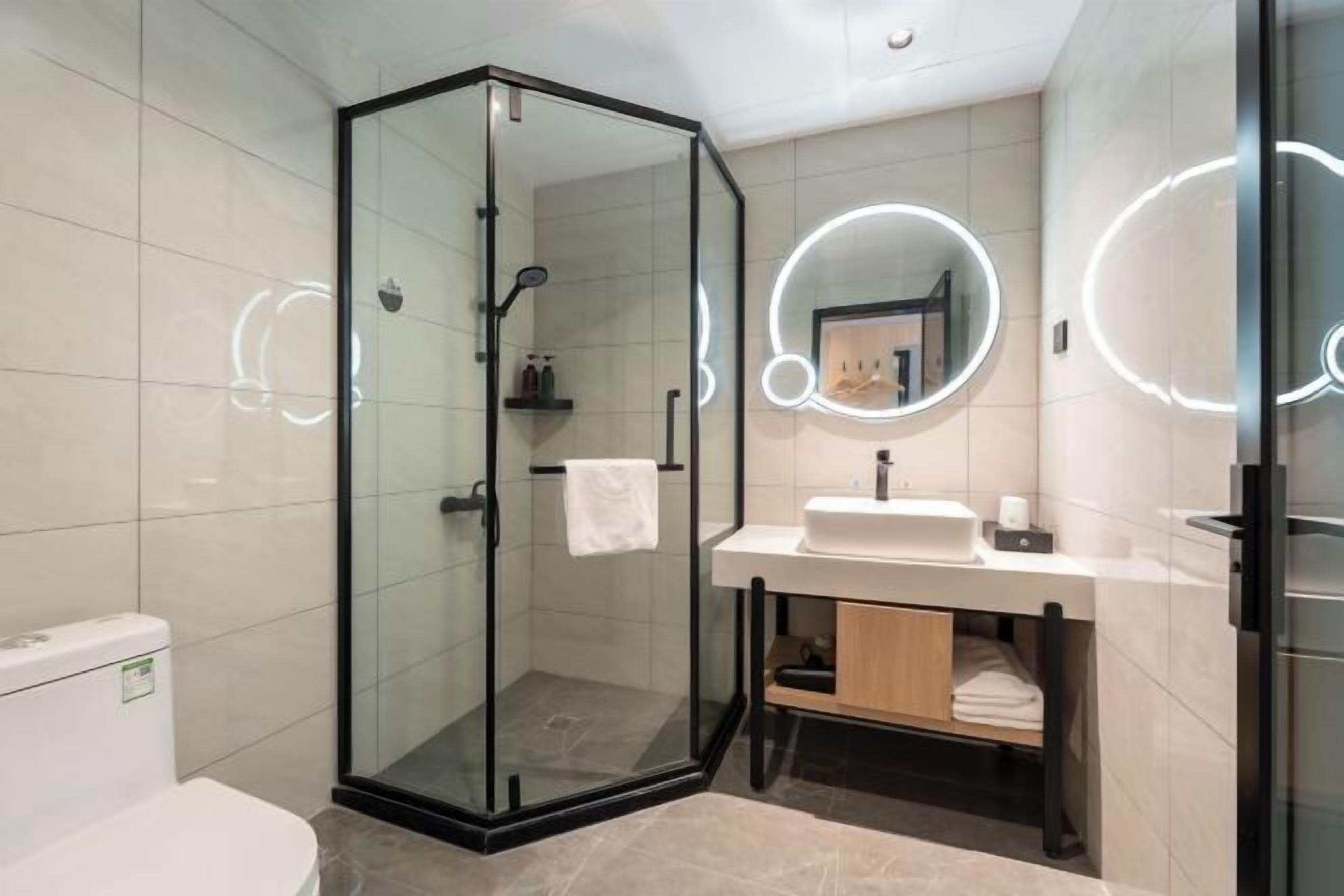Casa de banho moderna com chuveiro separado por vidro, lavatório branco e espelho com luzes circulares.