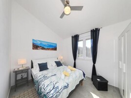 Quarto com cama, mesa de cabeceira, ventilador de teto, janela com cortinas e um quadro na parede.