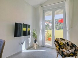 Quarto com televisão de tela plana na parede, um vaso com planta, uma cadeira com padrão floral e uma porta de vidro deslizante para área externa com um guarda-sol vermelho.