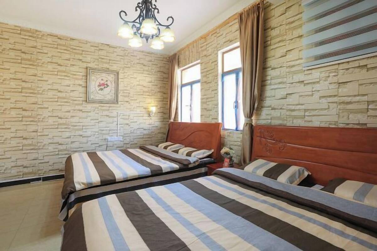 Quarto com parede de pedra, duas camas com roupa de cama listrada, cabeceira de madeira, lustre e janela com cortinas.