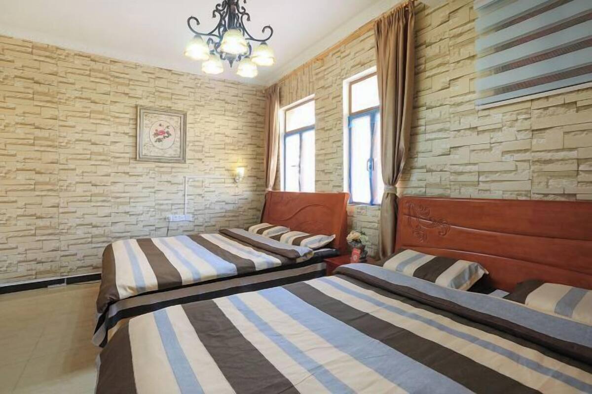 Quarto com parede de pedra, duas camas com roupa de cama listrada, cabeceira de madeira, lustre e janela com cortinas.
