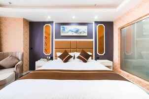 Quarto de hotel com uma cama grande, mesa de cabeceira, sofá e uma janela ampla.
