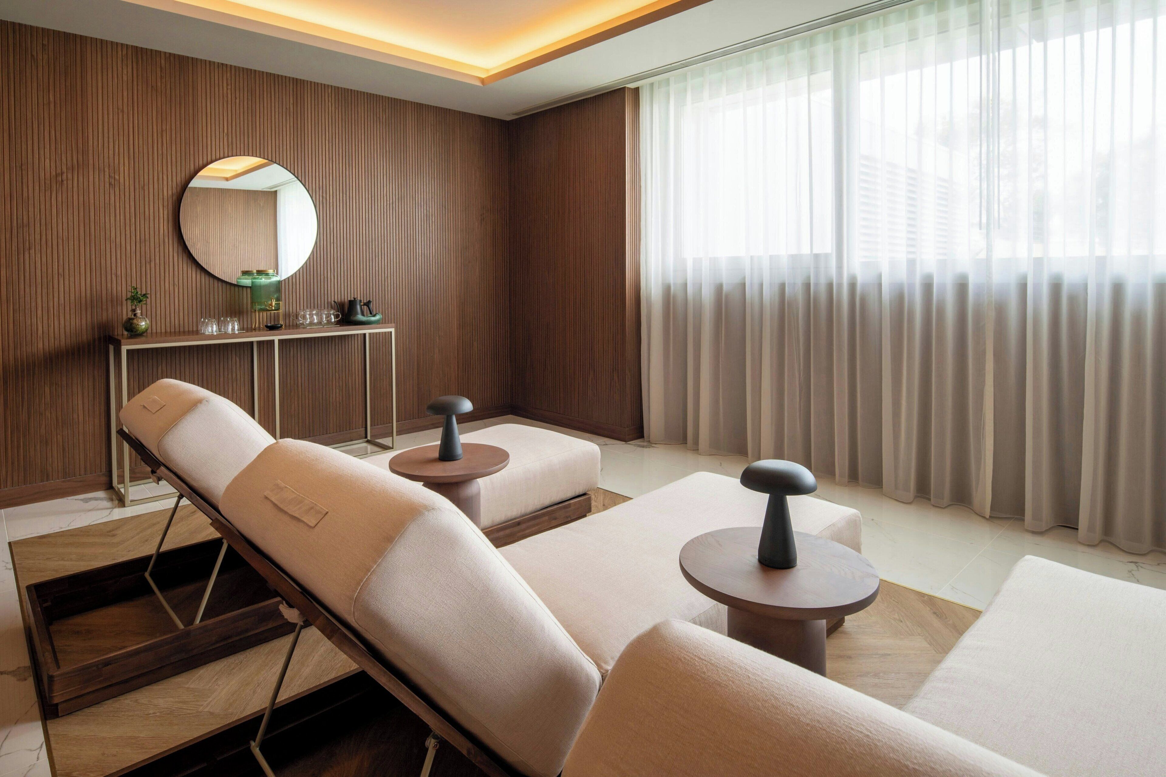 Um spa moderno com mesa de massagem, duas poltronas acolchoadas, um espelho redondo e um pequeno console com objetos decorativos.