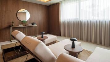 Um spa moderno com mesa de massagem, duas poltronas acolchoadas, um espelho redondo e um pequeno console com objetos decorativos.