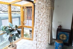 Quarto com uma grande janela, um vaso de planta, um tambor decorativo e uma pequena mesa com um objeto decorativo.
