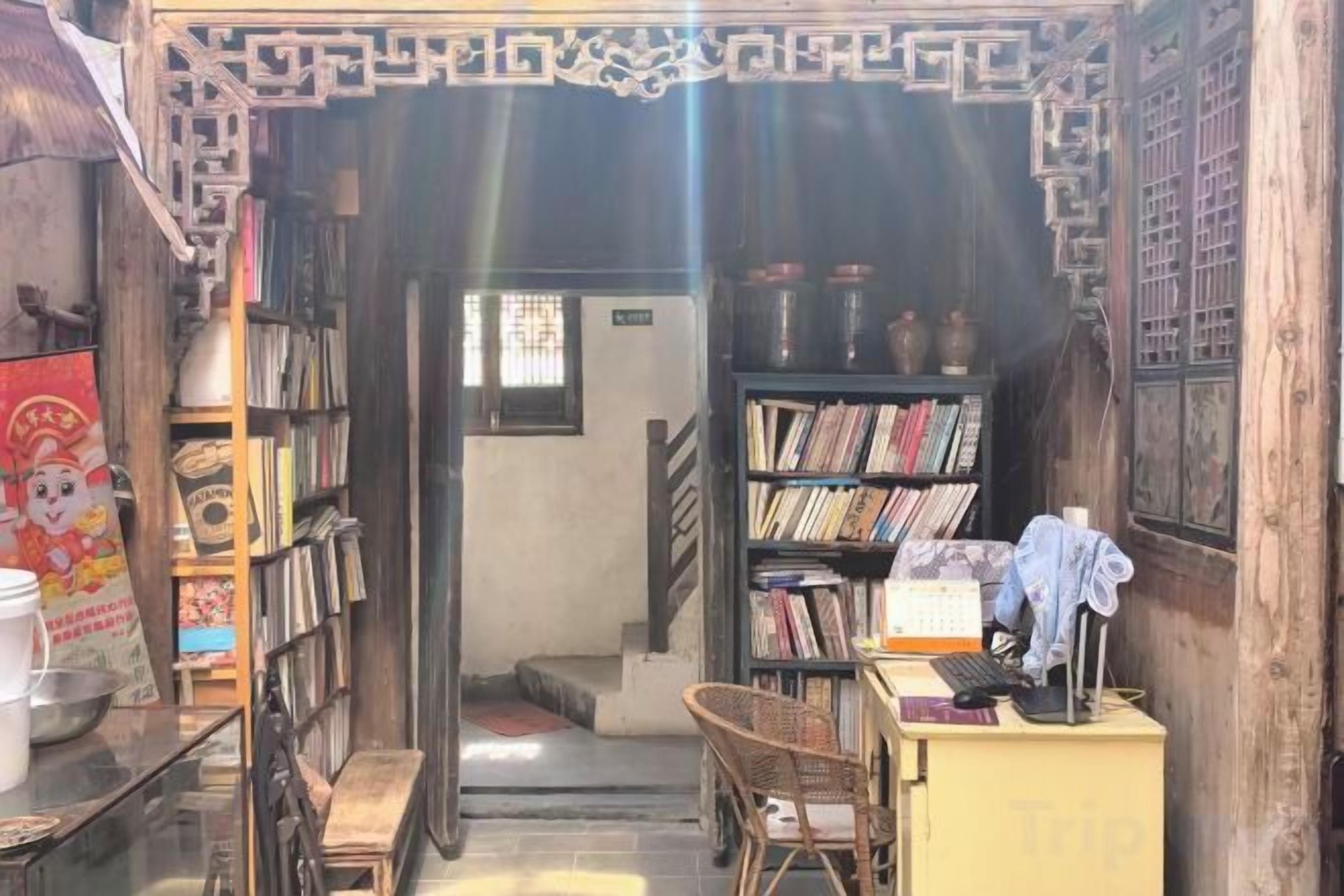 Um corredor estreito com estantes de livros em ambos os lados, uma escrivaninha de madeira com uma cadeira e uma pequena escadaria que leva a um andar superior.