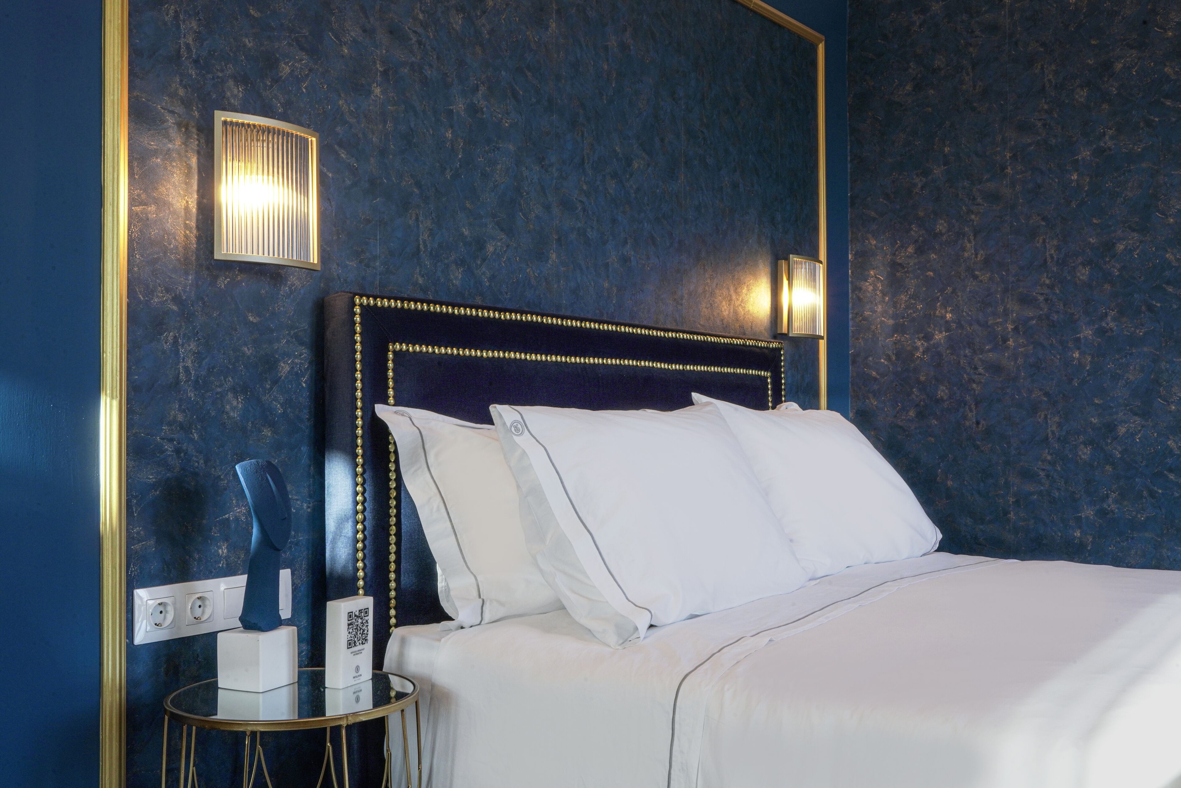 Quarto de hotel com parede azul escura texturizada, cama branca com cabeceira de madeira, duas luminárias de cabeceira e uma pequena mesa de cabeceira com uma luminária e um objeto decorativo.