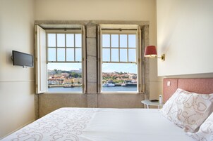 Quarto de hotel com cama, televisão, luminária e vista da cidade através de duas janelas.