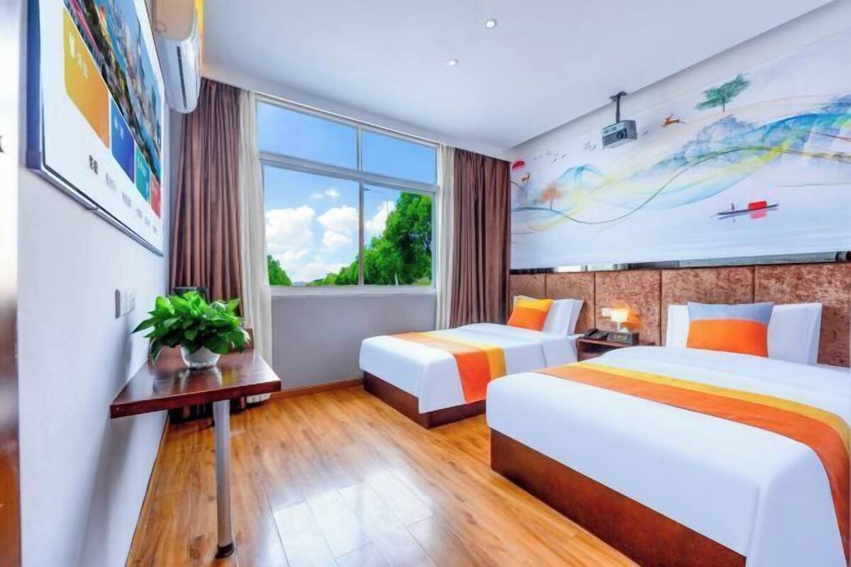 Quarto de hotel com duas camas, uma escrivaninha, um vaso de plantas, uma janela com vista e um mural na parede.