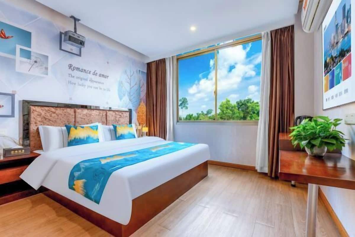 Quarto de hotel com uma cama grande, uma escrivaninha, vista para o exterior e uma parede com obras de arte e um projetor.