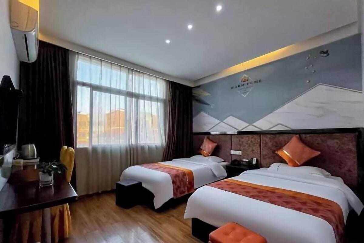 Quarto de hotel com duas camas, uma escrivaninha, uma cadeira e uma janela grande com cortinas.