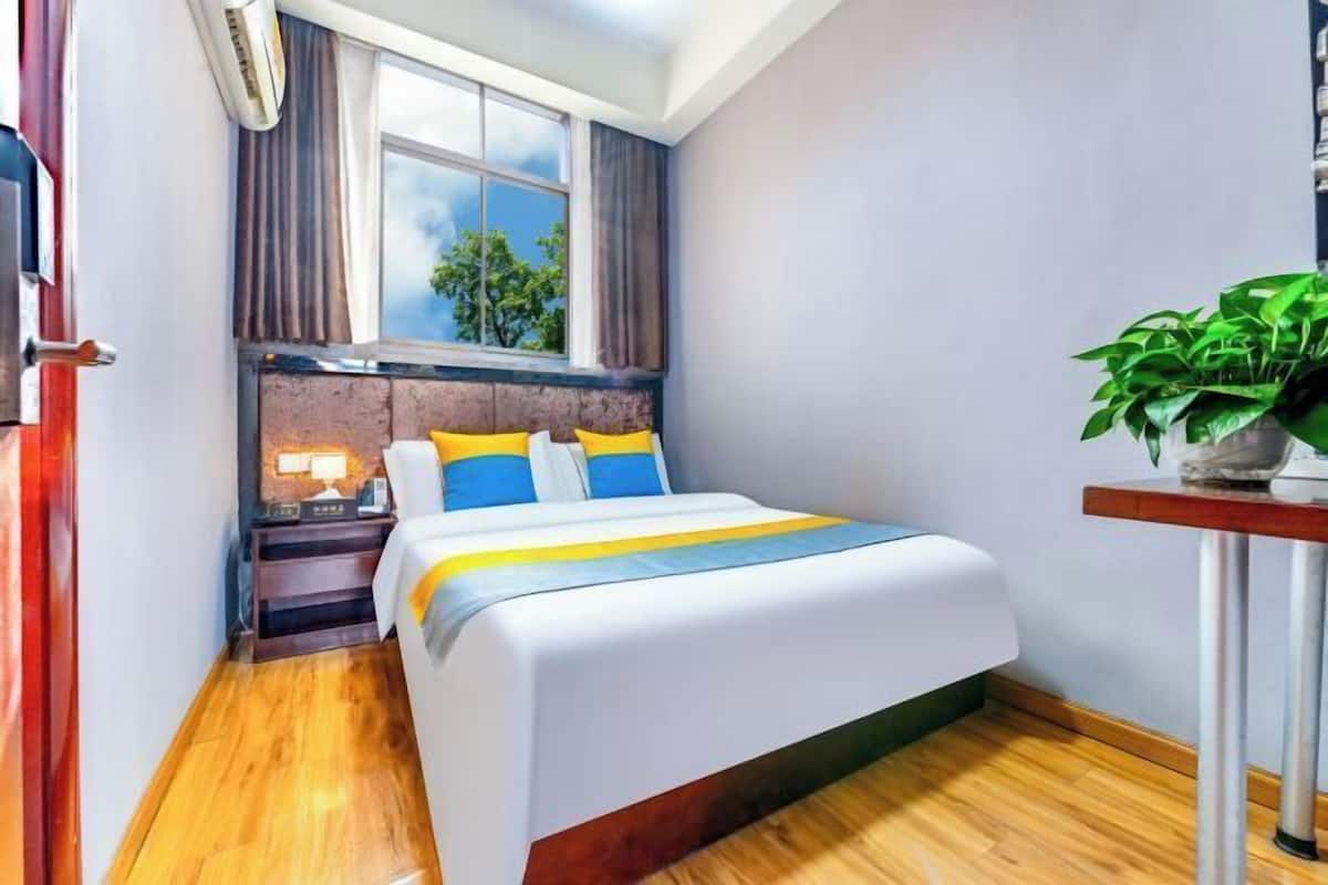 Quarto de hotel com cama, mesa de cabeceira, luminária, janela com cortinas, planta e piso de madeira.