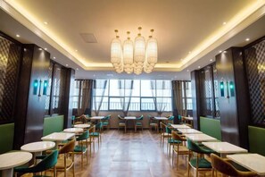 Restaurante com mesas e cadeiras redondas, um candelabro e janelas amplas.