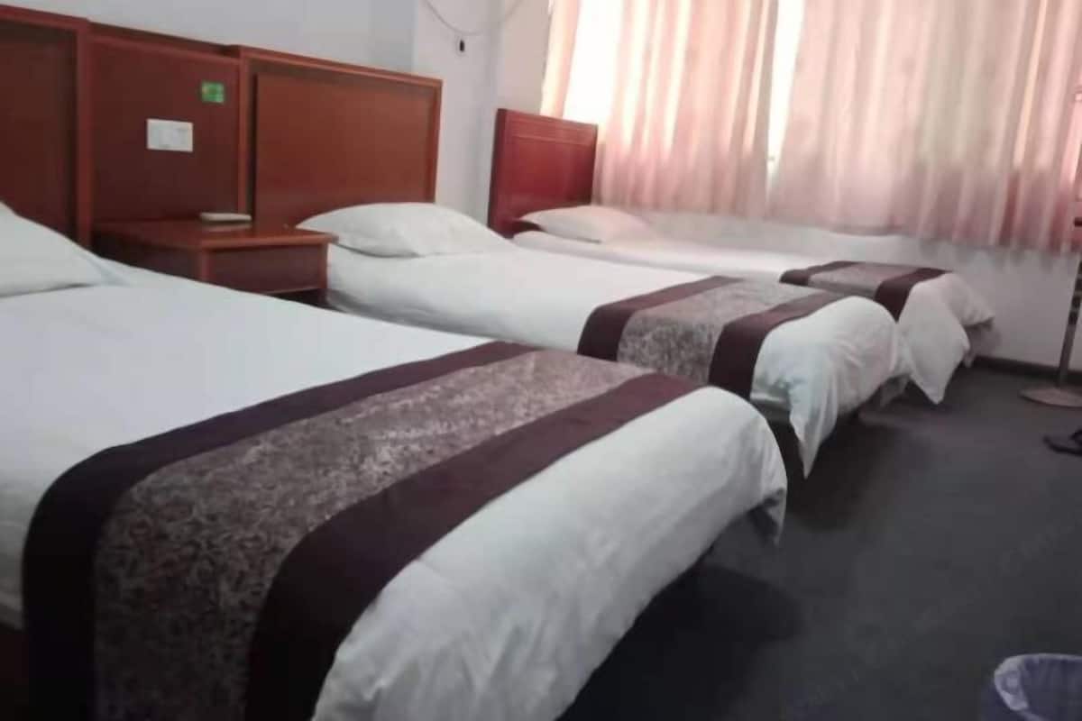 Quarto de hotel com três camas de solteiro, cabeceiras de madeira, pequenas mesas de cabeceira com luminárias e uma janela com cortinas.