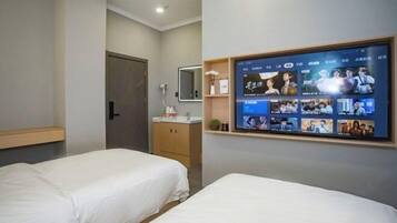 Quarto de hotel com duas camas, uma televisão fixada na parede exibindo diversos programas, um pequeno estante com artigos de higiene e uma porta que dá para outro cômodo.